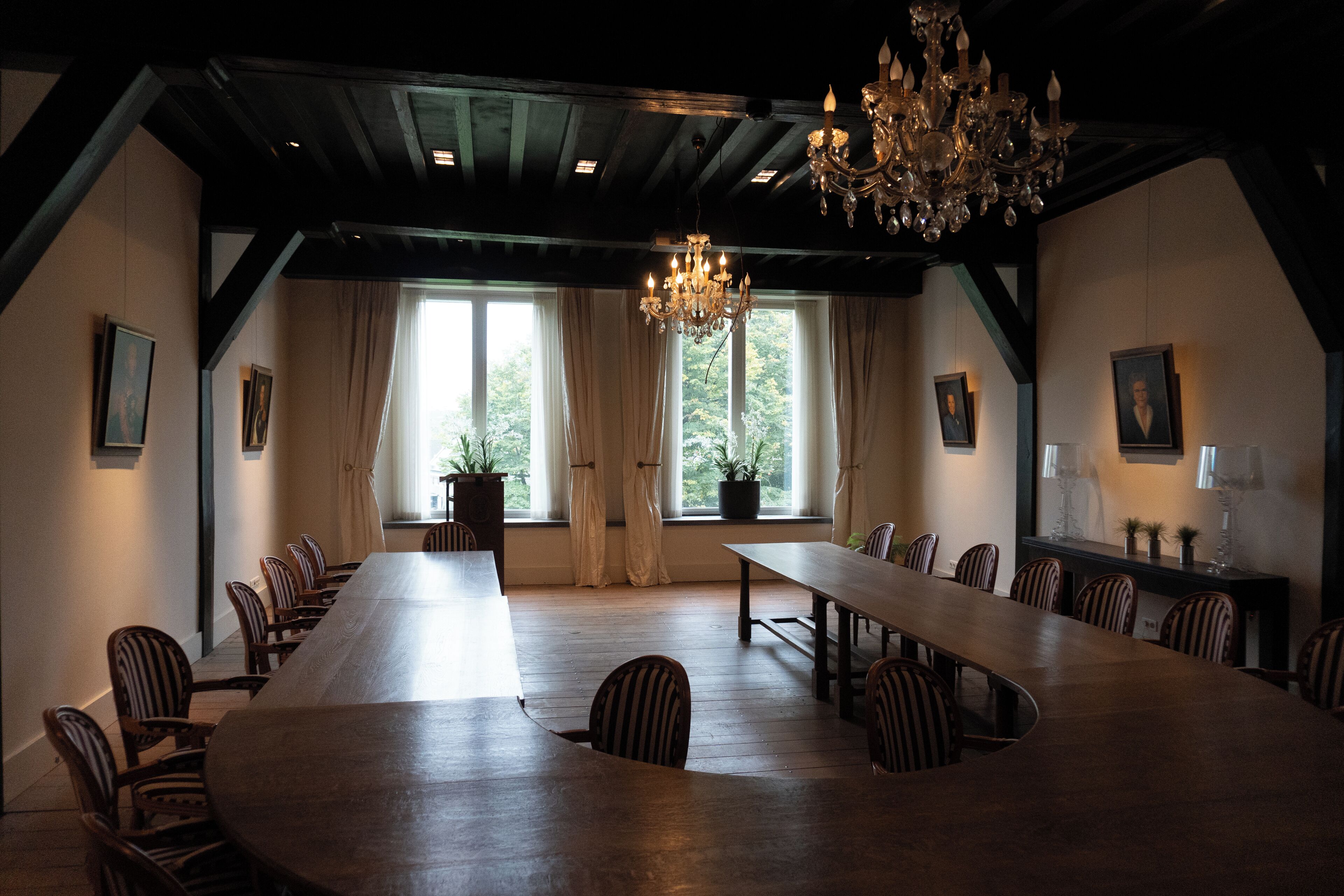 Uma sala de conferências com uma mesa comprida, cadeiras, um candelabro e grandes janelas com cortinas.