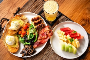 Prato de pequeno-almoço com ovos, bacon, cogumelos e tomate em torradas, um prato de fruta e um copo de sumo.