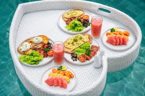 Disposição de mesa para refeição com pratos de comida, um copo de suco e saleiros e pimenteiros sobre uma bandeja de vime branca, perto de uma piscina.