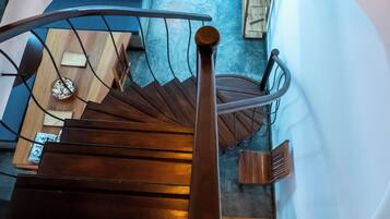 Uma escadaria em espiral com corrimão de madeira, que leva a um quarto com piso azul texturizado e uma mesa de madeira.