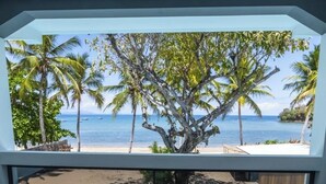Quarto com uma grande janela que dá para uma praia com palmeiras e o oceano.