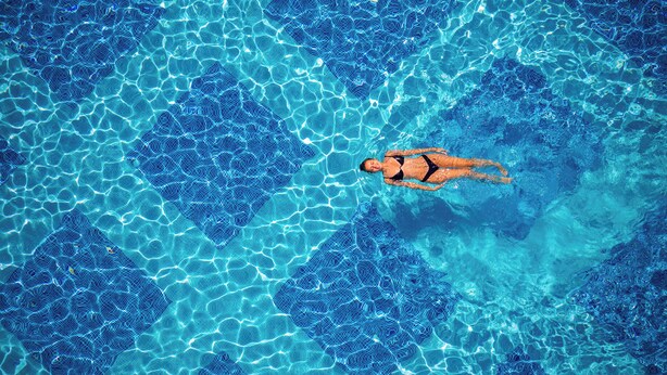Uma pessoa nadando em uma piscina azul cristalina com um padrão geométrico de azulejos.