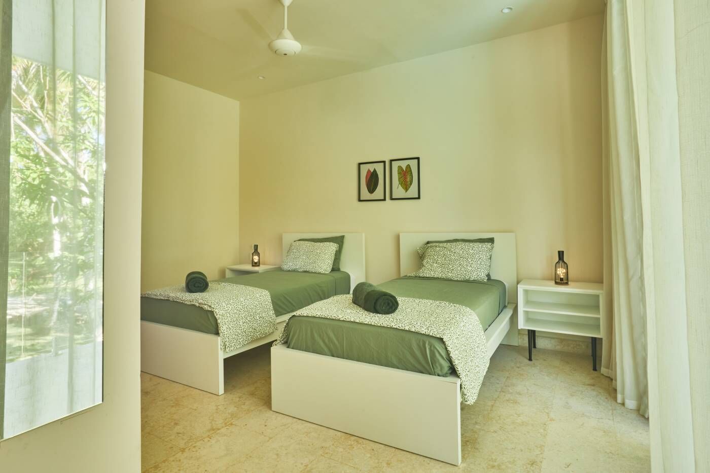 Quarto com duas camas, um ventilador de teto, mesa de cabeceira e janela com cortinas.