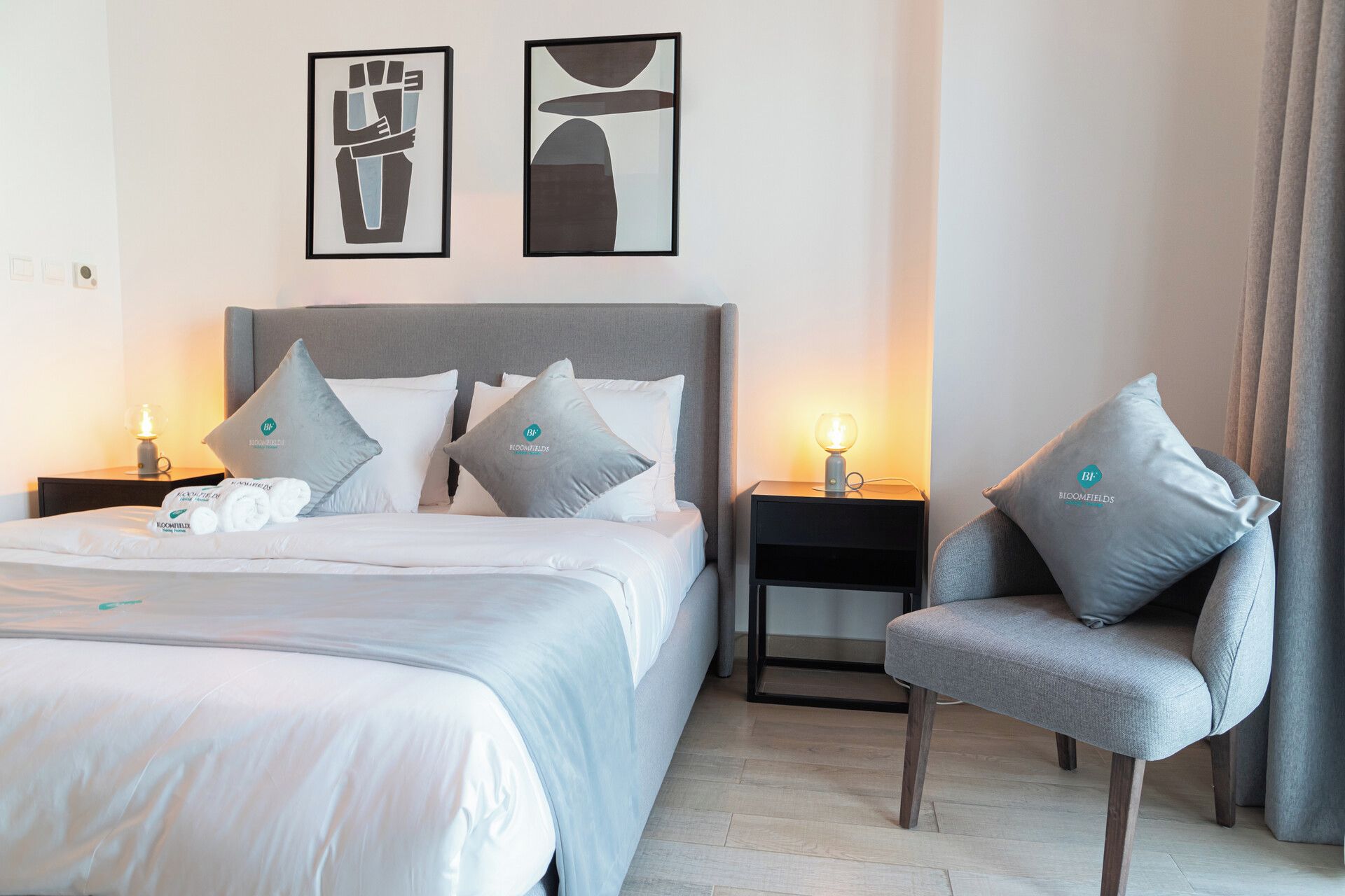Quarto de hotel com uma cama, uma cadeira, duas mesas de cabeceira e duas obras de arte na parede.