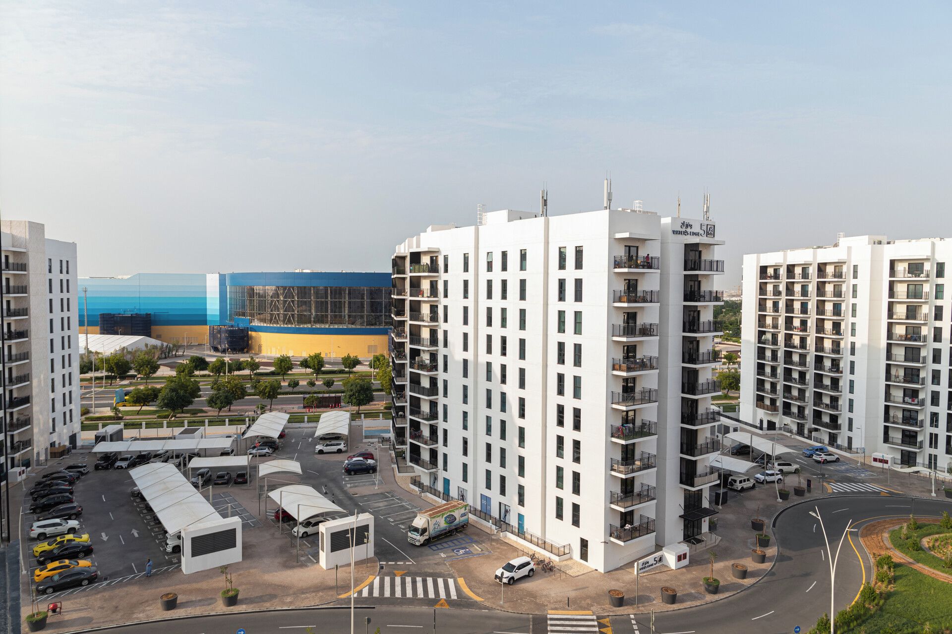 Um edifício moderno de muitos andares com várias varandas, área de estacionamento e um edifício azul ao fundo.