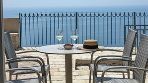 Mesa redonda com cadeiras de vime, uma tigela de frutas e dois copos de vinho em uma varanda com vista para o mar.