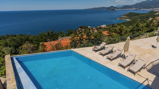 Piscina retangular com espreguiçadeiras e guarda-sóis, rodeada por um muro de pedra e com vista para o mar e colinas.