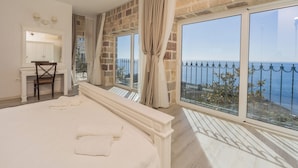 Quarto com uma cama grande, uma escrivaninha e vista para o mar através de amplas janelas.