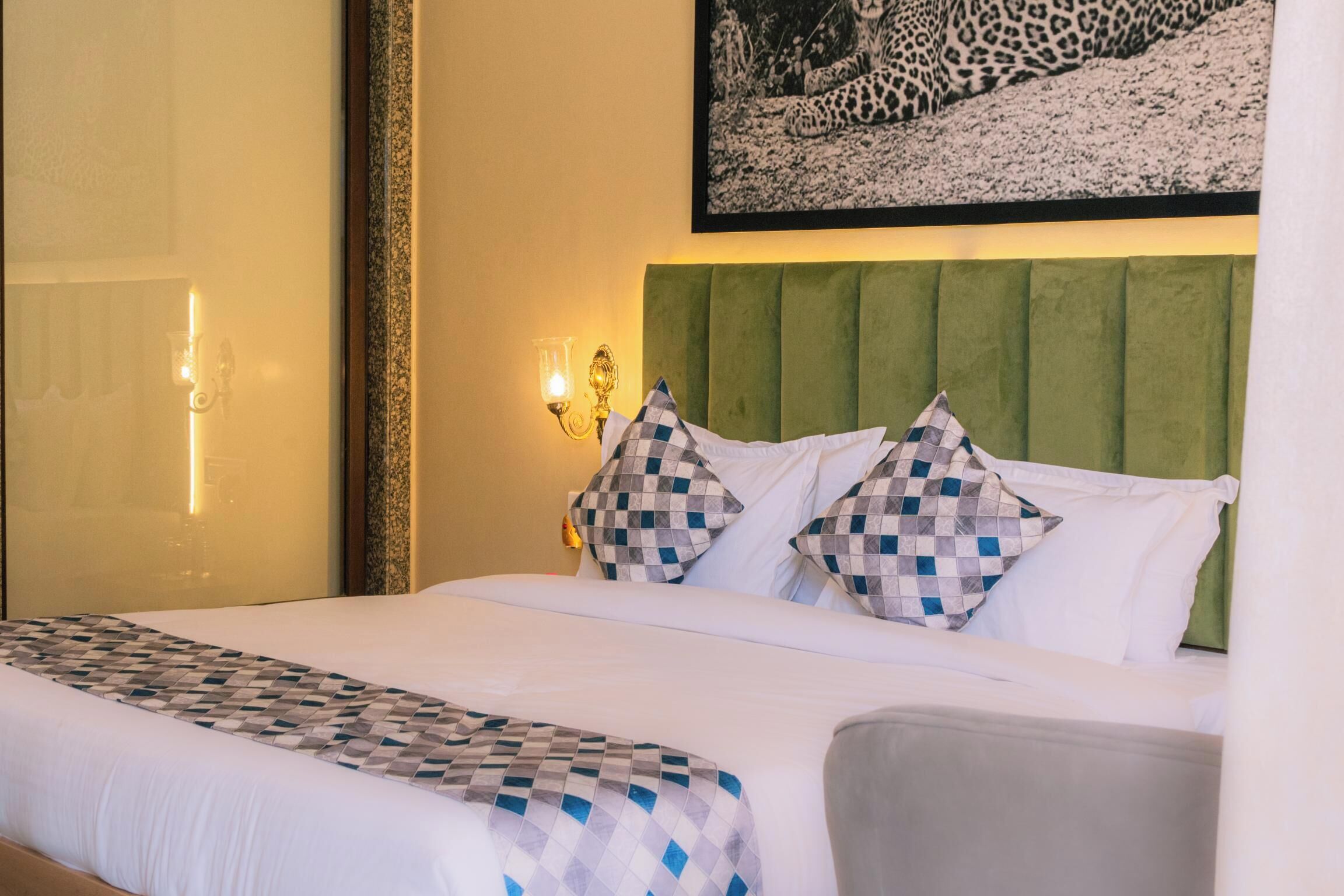 Quarto de hotel com uma cama grande, manta xadrez, cabeceira estofada verde e uma obra de arte com estampa de leopardo acima da cama.