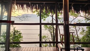 Quarto com deck de madeira, telhado de palha e vista para um corpo d'água.