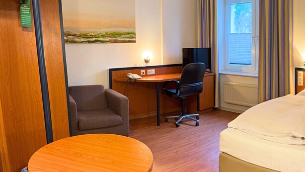 Quarto de hotel com uma cama, uma secretária com cadeira, uma mesa redonda pequena, uma janela com cortinas e um quadro na parede.