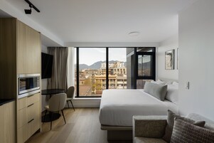 Quarto de hotel moderno com uma cama grande, uma mesa de escritório e vista para a cidade através de janelas do chão ao teto.