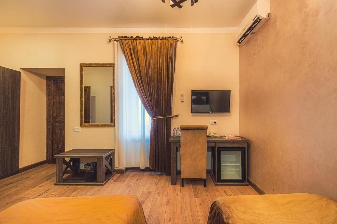Quarto de hotel com piso de madeira, uma escrivaninha com cadeira, cama, televisão e janela com cortinas.