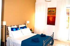 Quarto de hotel com cama, cortinas brancas, um quadro na parede e mesa de cabeceira com abajur e flor.