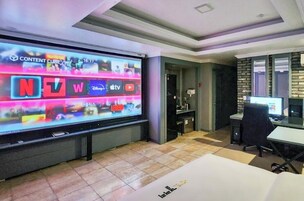 Quarto moderno com uma grande TV de tela plana exibindo diversos serviços de streaming. Há uma mesa com um computador e uma cadeira. O quarto possui piso cerâmico e uma parede de destaque em tijolo.