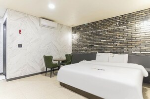 Quarto de hotel com cama, uma pequena mesa, duas cadeiras, parede de mármore, parede de tijolos com uma frase motivacional e um aparelho de ar condicionado.