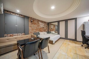 Quarto de hotel moderno com parede de destaque em tijolo, uma cama grande, um sofá, uma mesa de jantar com cadeiras e uma escrivaninha com cadeira.