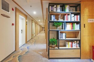 Um corredor de hotel com piso em carpete, uma estante com livros e plantas, e portas dando acesso aos quartos.