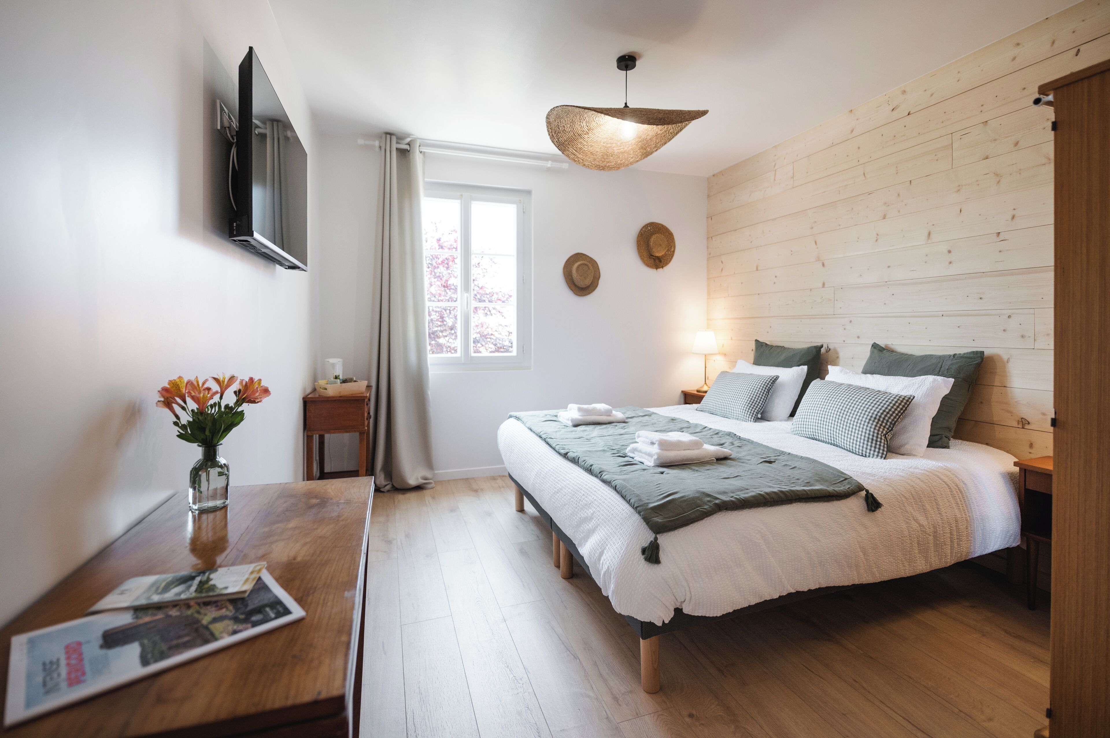Quarto com cama, mesas de cabeceira, TV fixada na parede, janela com cortinas e uma parede de madeira.