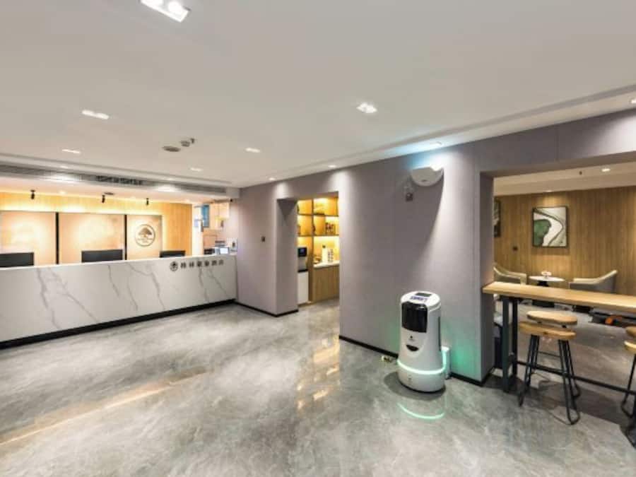 Uma área de recepção de hotel com balcão de mármore, um quiosque de autoatendimento e um bar com banquetas.