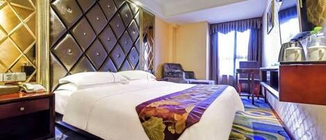 Quarto de hotel com cama grande, televisão, escrivaninha com telefone e janela com cortinas.