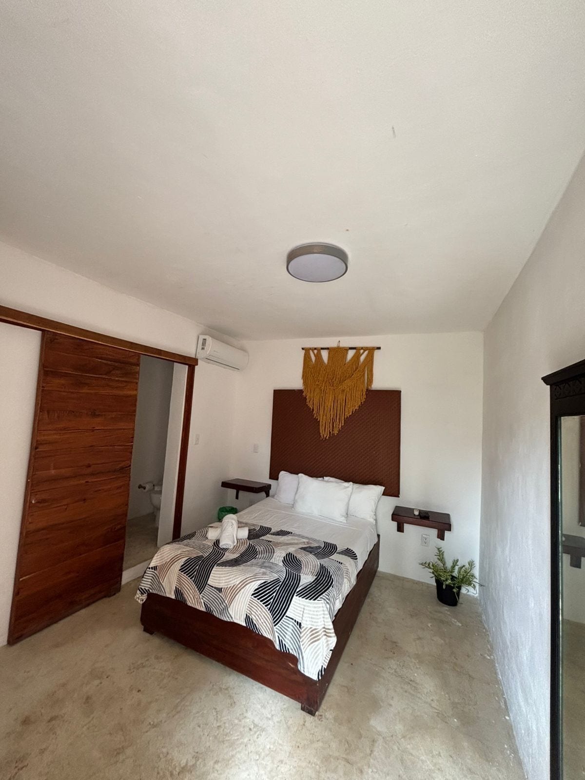 Quarto com cama, guarda-roupa de madeira, mesas de cabeceira, decoração de macramé pendurada na parede e um vaso com planta.