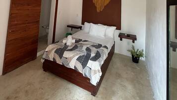 Quarto com cama, guarda-roupa de madeira, mesas de cabeceira, decoração de macramé pendurada na parede e um vaso com planta.