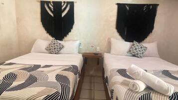 Quarto com duas camas, cada uma com colcha e almofadas estampadas. Há uma pequena mesa de cabeceira com um abajur e um telefone.