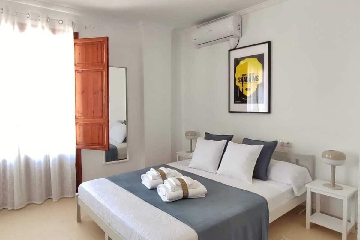 Quarto com cama, duas mesas de cabeceira, ar-condicionado fixado na parede, quadro emoldurado e janela com cortinas.