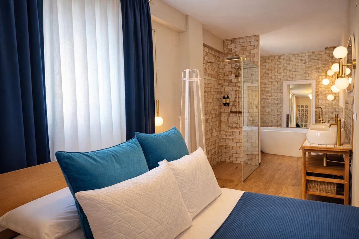 Quarto com cama, mesa de cabeceira, chuveiro, banheiro com espelho e parede de tijolos.