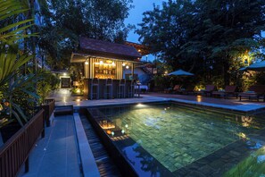 Uma área de piscina com bar e assentos, cercada por árvores e um edifício com telhado em azulejos.