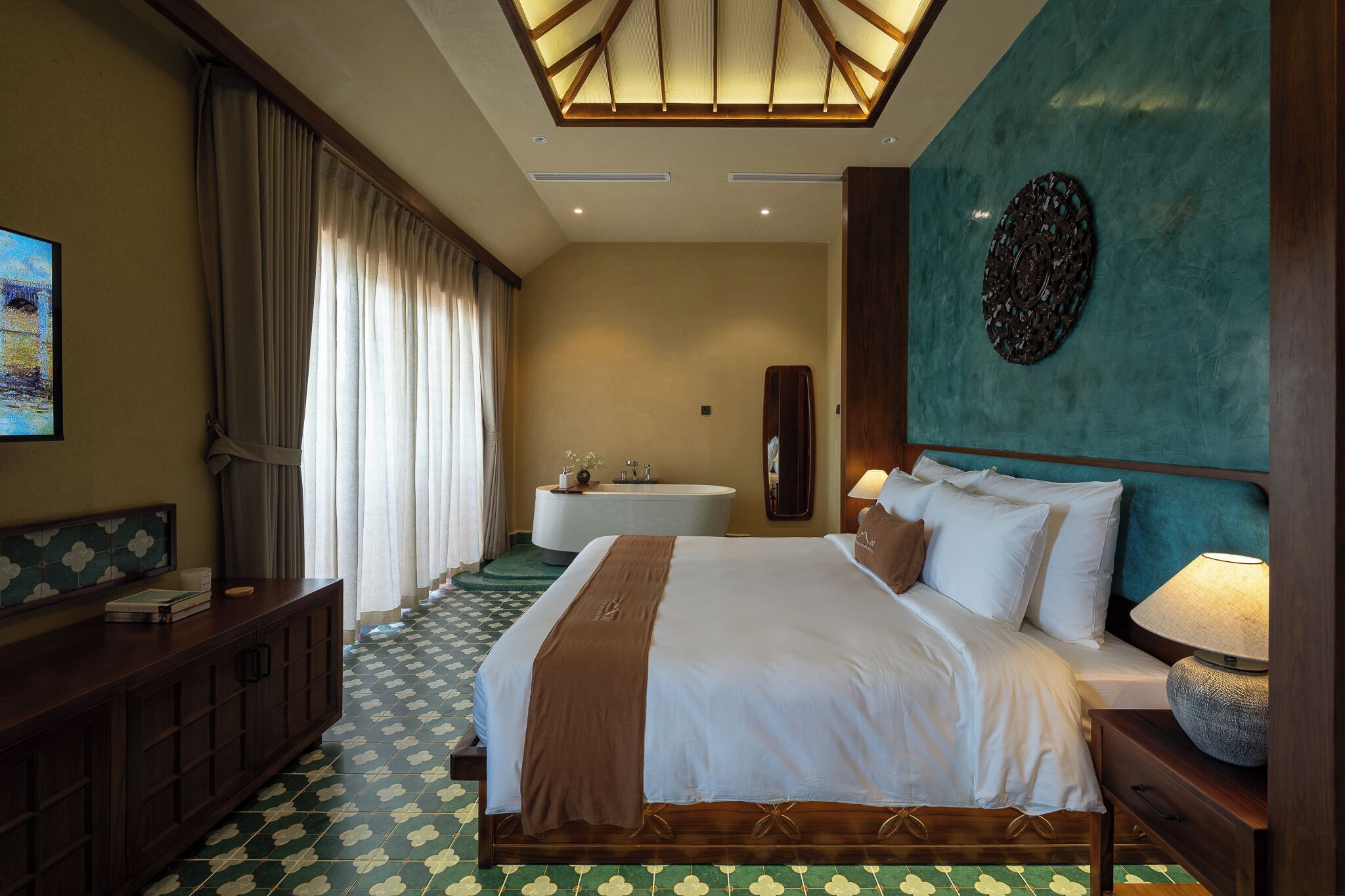 Quarto de hotel com uma cama grande, mesa de cabeceira com luminária, penteadeira com espelho e janela com cortinas.