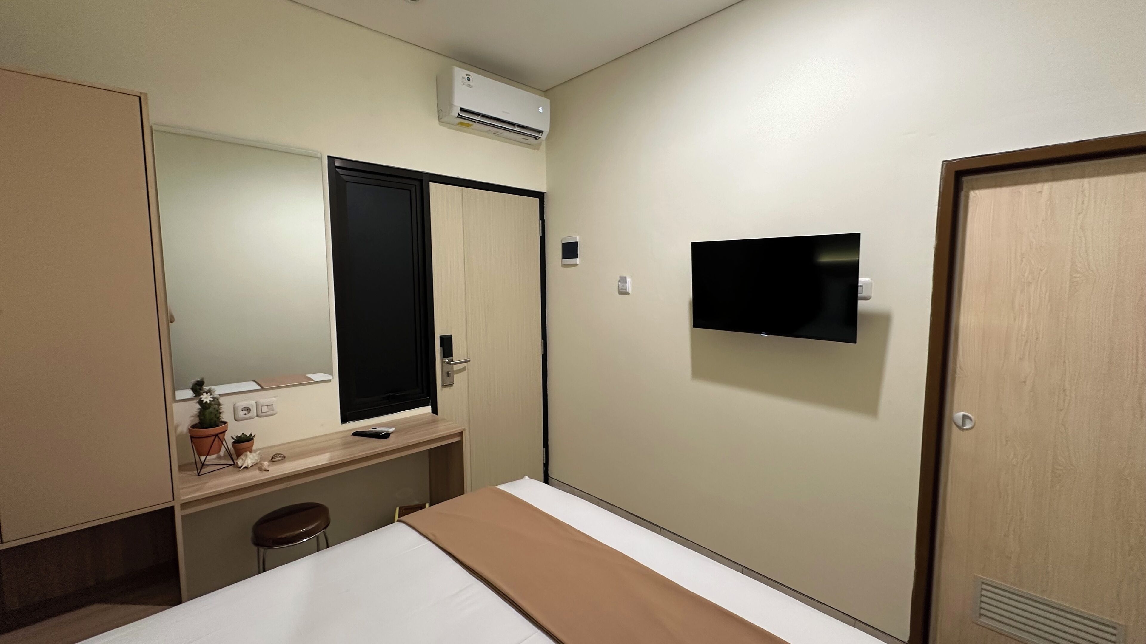 Quarto de hotel com cama, escrivaninha e cadeira, espelho, televisão na parede e uma porta.