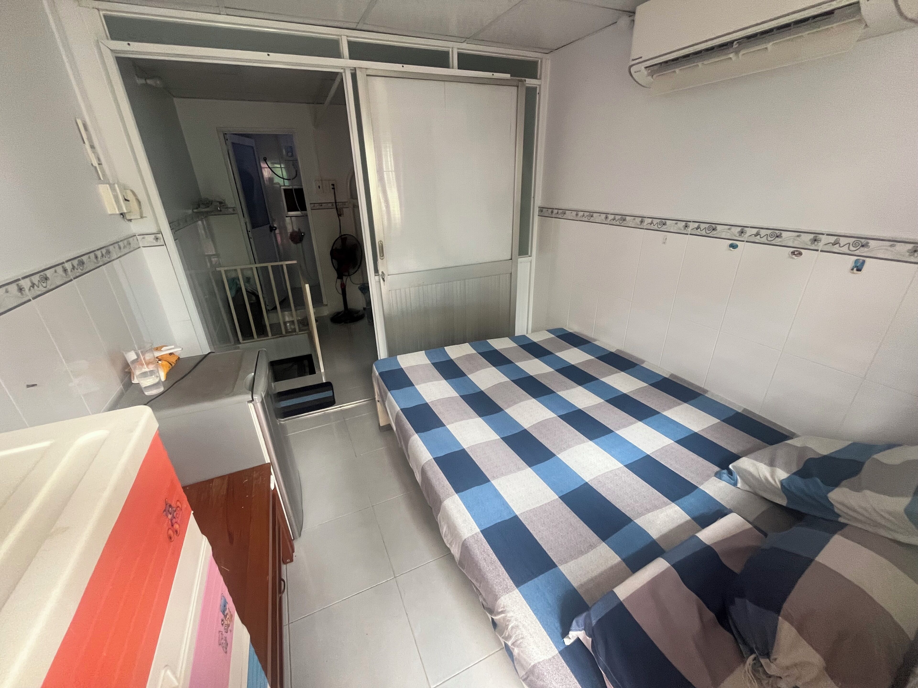 Quarto pequeno e compacto com cama, escrivaninha e armário. Há uma porta que dá para outro espaço e um radiador na parede.