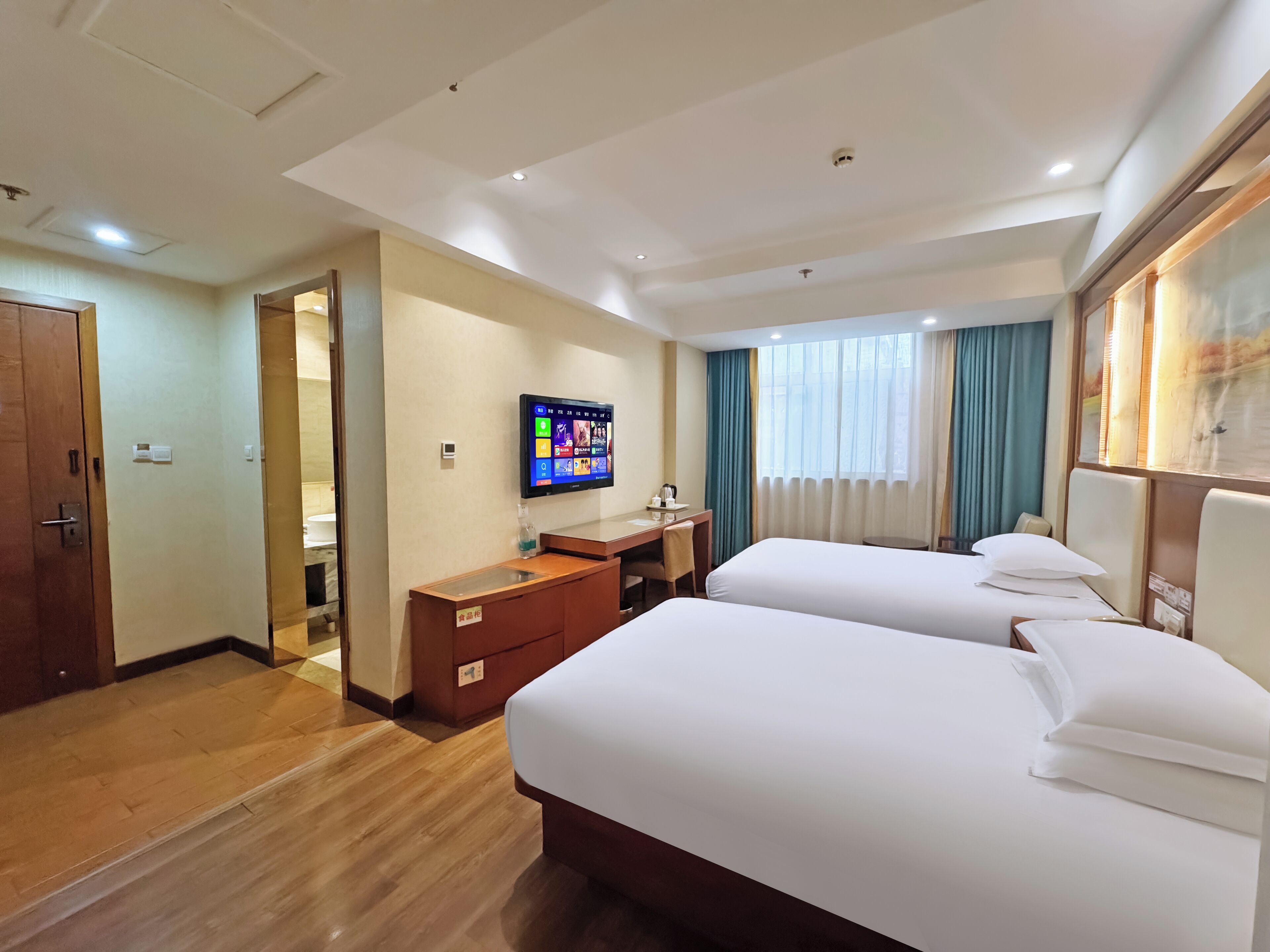 Quarto de hotel com duas camas, uma escrivaninha, uma televisão e uma janela com cortinas.