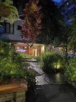 Vista noturna de um pátio de hotel com um elemento aquático, vegetação exuberante e uma entrada bem iluminada.