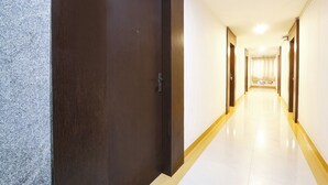Um corredor longo e estreito, com uma porta de madeira à esquerda e um chão claro revestido de azulejos.