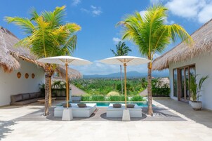 Área de piscina com duas espreguiçadeiras e guarda-sóis, cercada por palmeiras e vista para uma paisagem montanhosa.
