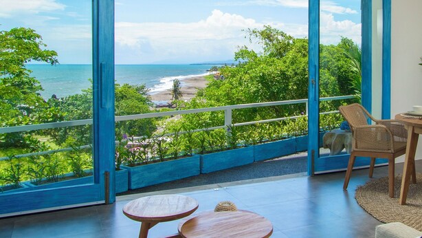 Varanda com vista para o oceano e a praia, móveis de madeira e guarda-corpo azul.
