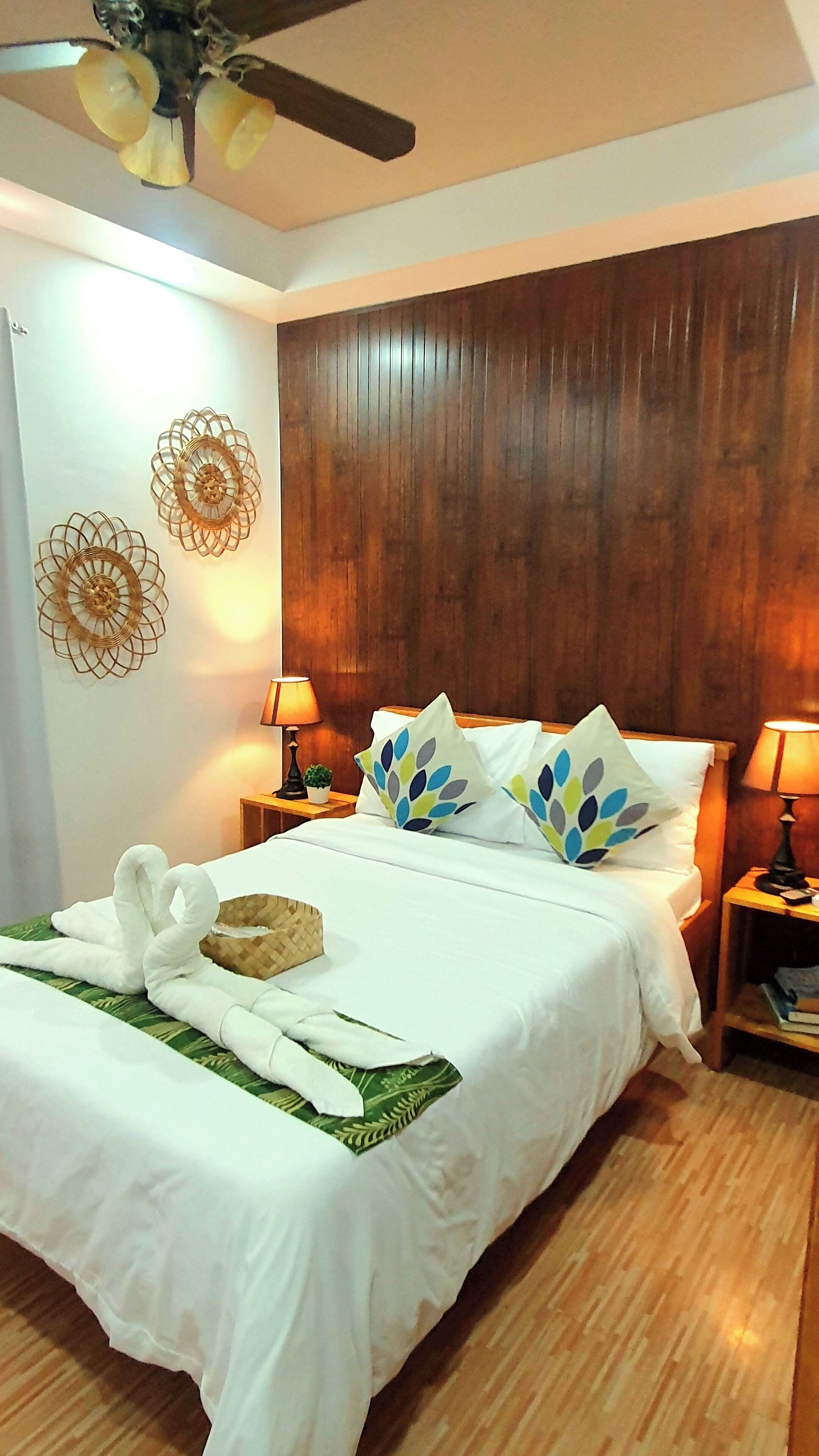 Uma cama bem arrumada com lençóis brancos e almofadas decorativas, duas luminárias de cabeceira, um ventilador de teto e um painel de cabeceira de madeira.
