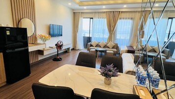 Quarto de hotel moderno com mesa de jantar, cadeiras, TV, sofá e varanda com cortinas.