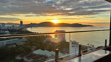 Vista do pôr do sol a partir de uma varanda em um arranha-céu, com vista para uma paisagem urbana costeira, com edifícios, uma praia e uma ilha ao longe.