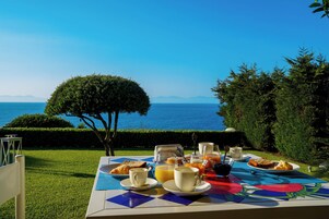Mesa posta para o café da manhã com vista para o mar, incluindo pratos de comida, xícaras e um copo de suco.