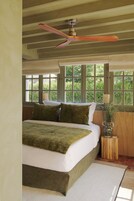Quarto com uma cama grande, um ventilador de teto e vista para o verde através de janelas amplas.
