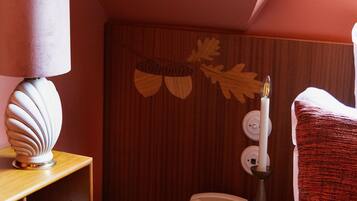 Quarto com prateleira de madeira contendo uma luminária, um telefone e uma caixa de lenços. Há uma cama com travesseiro vermelho e uma parede com detalhes em folhagens.