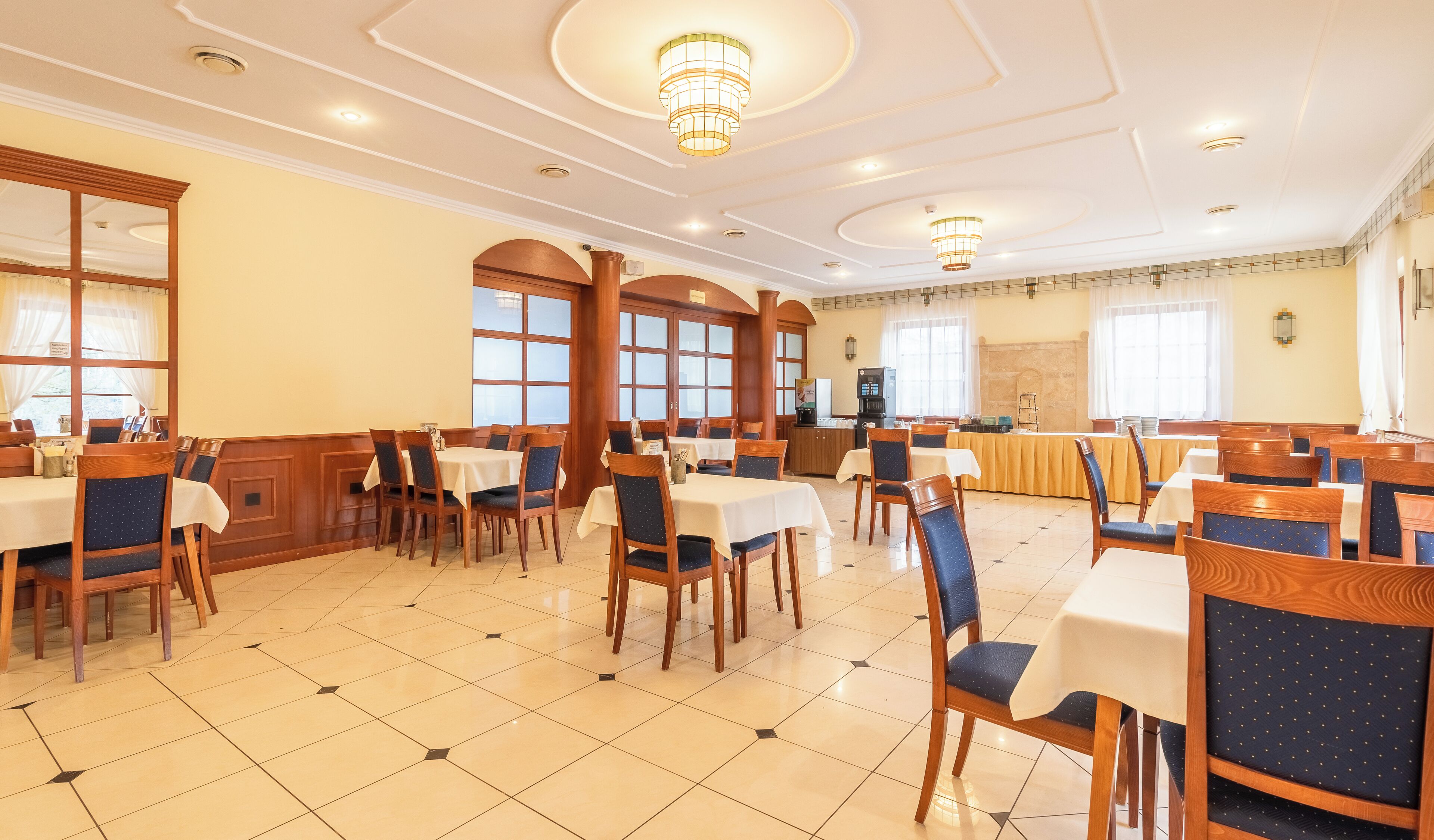 Uma ampla sala de jantar com várias mesas e cadeiras, grandes janelas e um lustre.