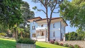 Um edifício de dois andares com varanda, rodeado por vegetação exuberante e plantas floridas.
