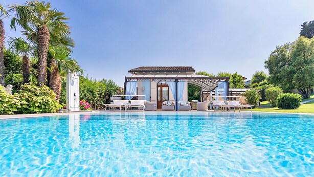 Piscina com espreguiçadeiras e um edifício com pérgola ao fundo.