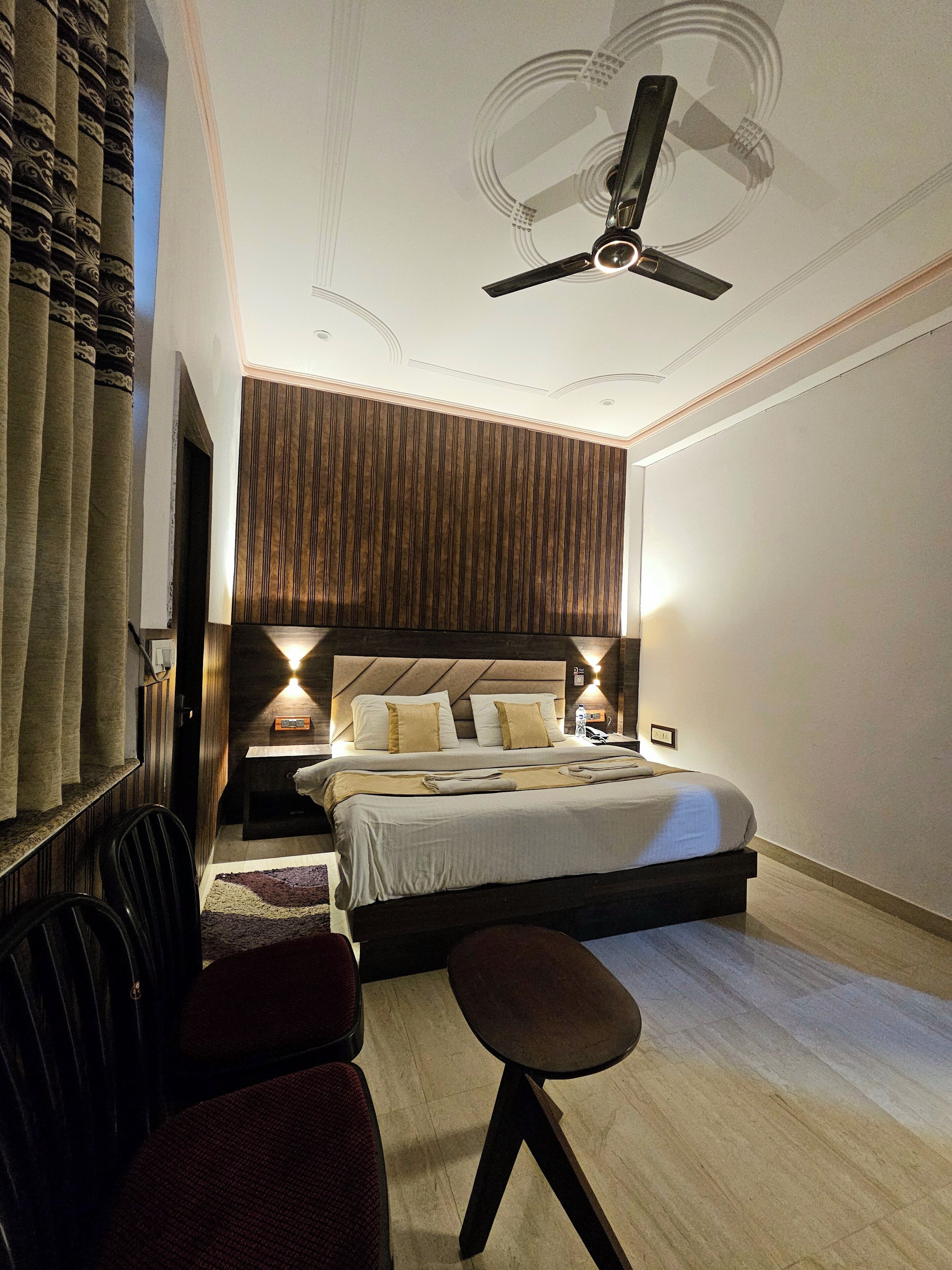 Quarto de hotel moderno com uma cama grande, um ventilador de teto, uma mesinha e um espaço de estar com uma poltrona.