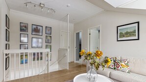Uma sala de estar moderna com um sofá branco, uma mesa de centro e um vaso com flores. Há quadros nas paredes e uma escada que leva a outro cômodo.